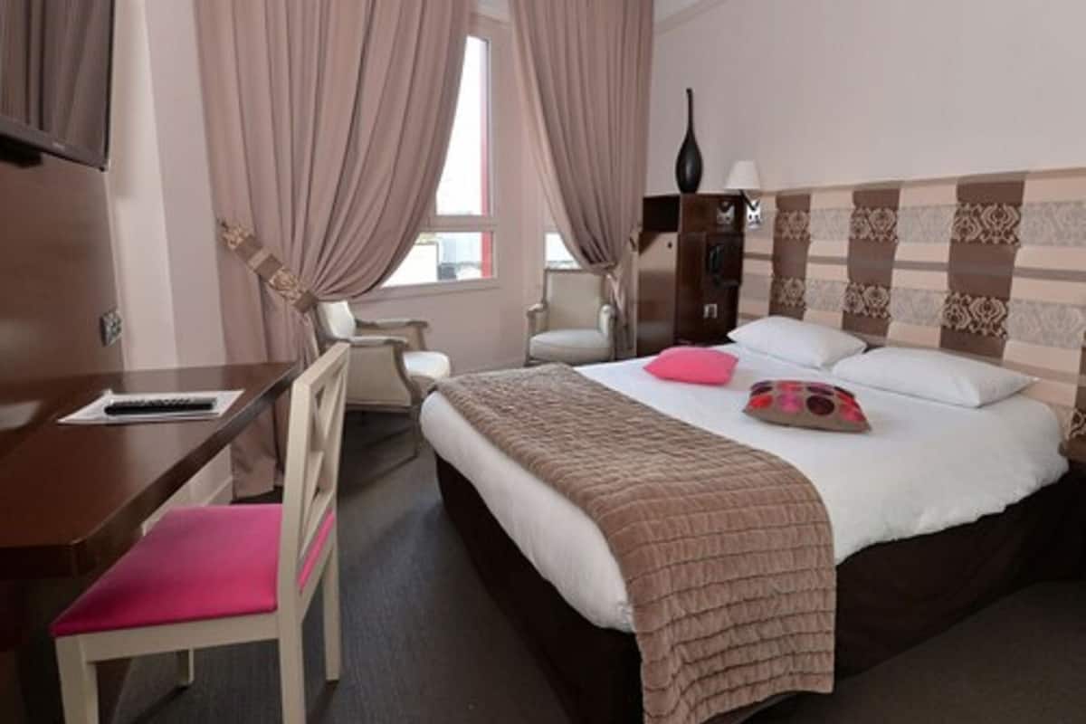 Quarto de hotel com uma cama, uma mesa, uma cadeira e uma janela com cortinas.
