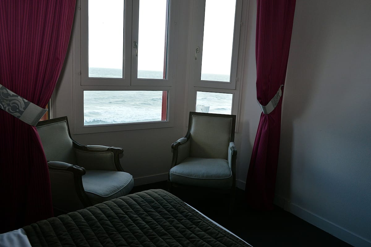 Quarto com cama, duas cadeiras e janela com vista para o mar.