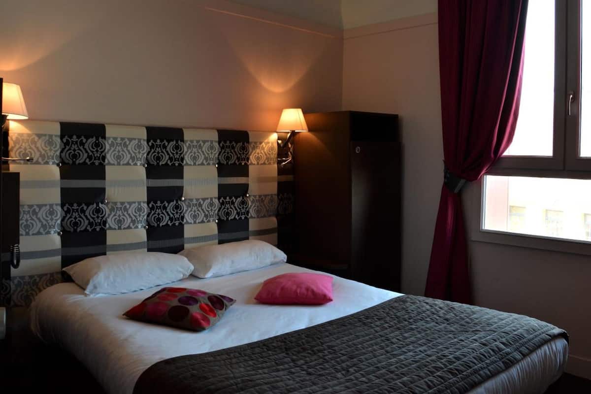 Quarto de hotel com cama, duas almofadas, manta acolchoada, cabeceira estampada, janela com cortinas e luminárias fixadas na parede.