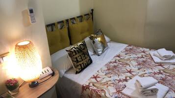 Uma cama bem arrumada com colcha floral, um abajur sobre a mesa de cabeceira e um telefone fixado na parede.