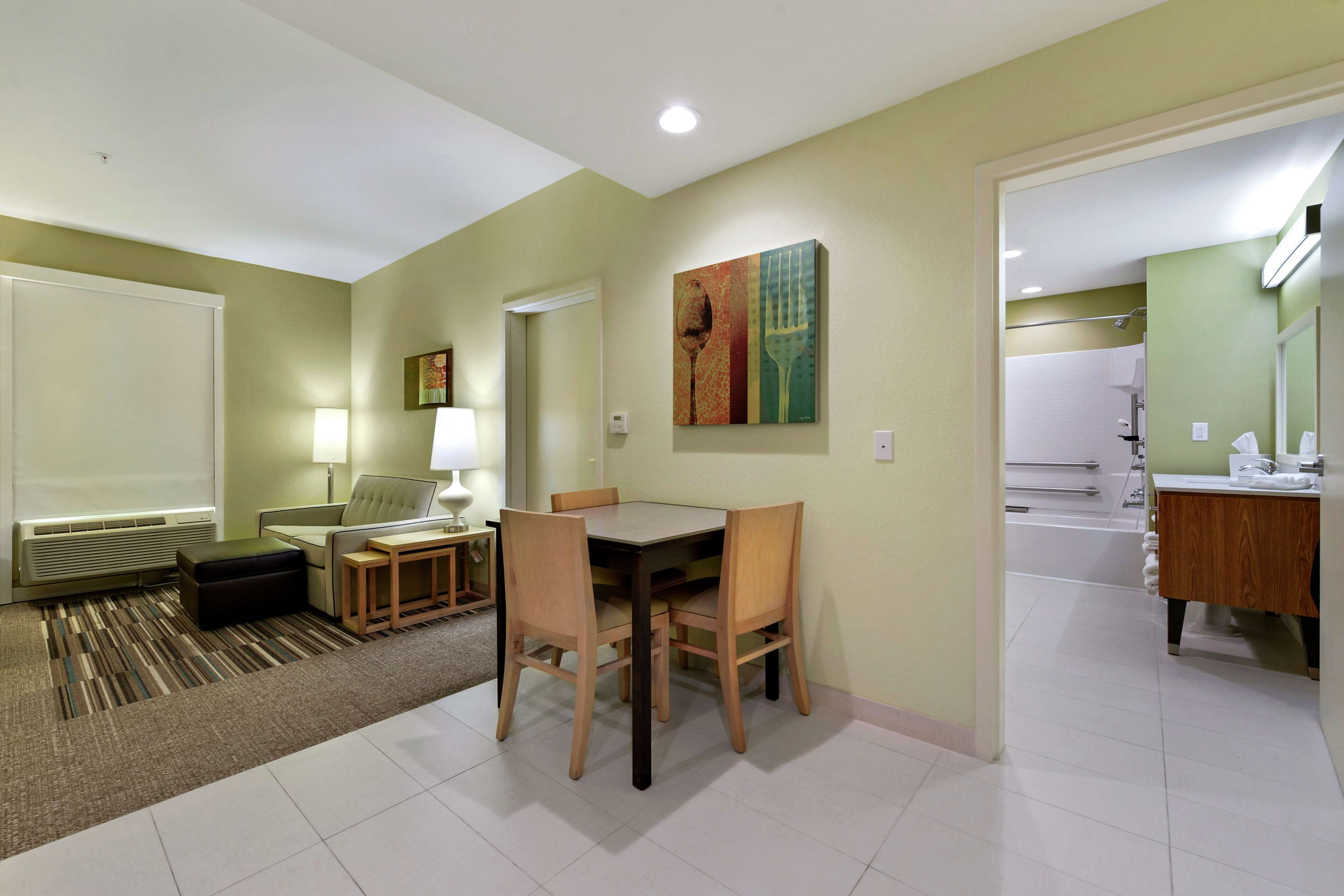 Quarto de hotel com área de jantar, sofá, luminária e pintura na parede.