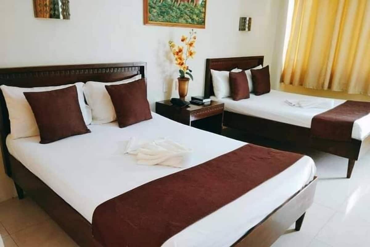 Quarto de hotel com duas camas, cada uma com cabeceira de madeira, roupa de cama branca e almofadas decorativas castanhas. Há uma mesinha de cabeceira com um vaso de flores e uma luminária entre as camas.