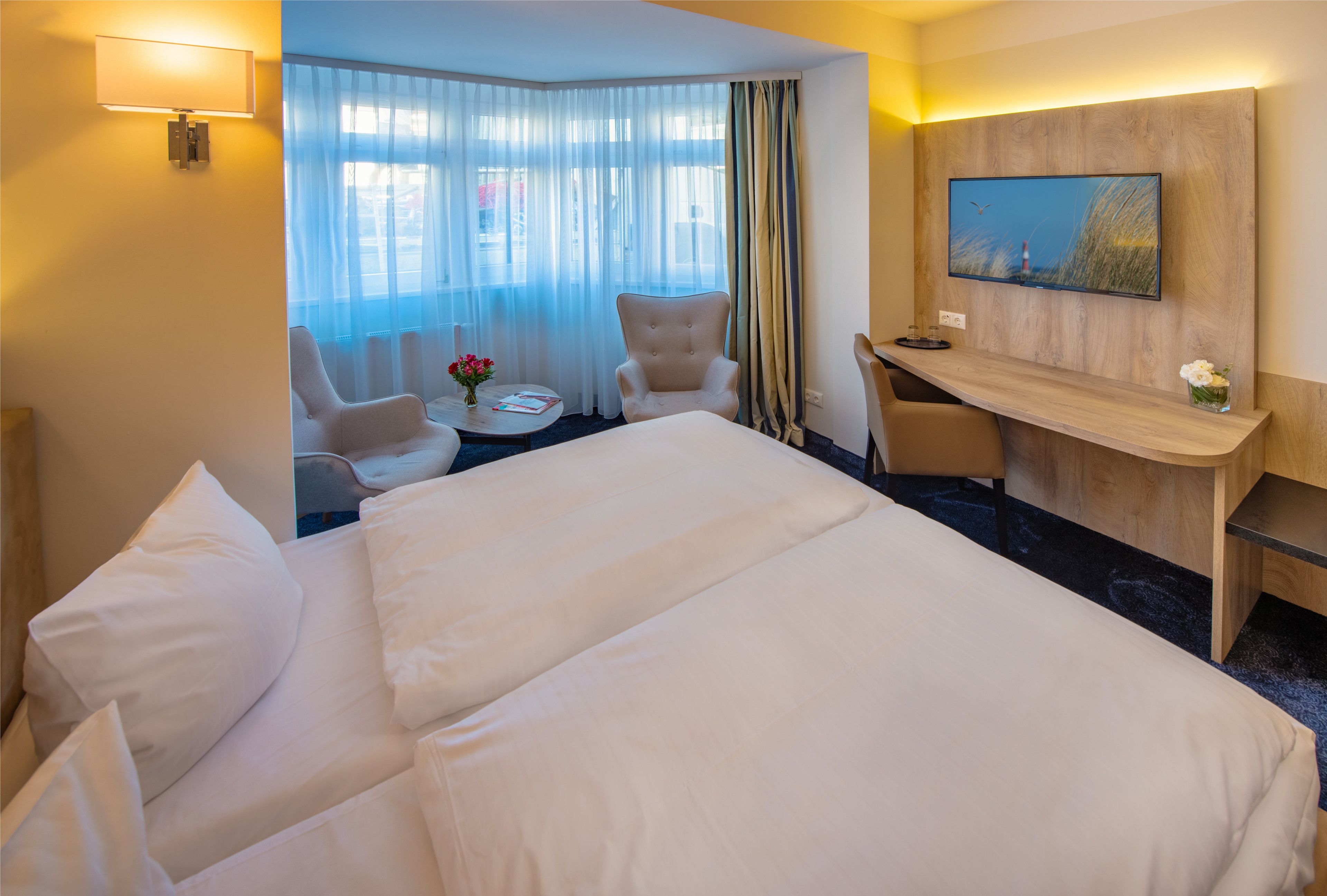 Quarto de hotel com uma cama grande, uma escrivaninha, dois poltronas, uma mesa pequena com um vaso de flores e um quadro na parede.