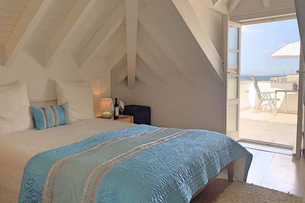 Quarto com uma cama grande, uma mesinha de cabeceira, uma cadeira e vista para o mar através de uma porta de vidro.