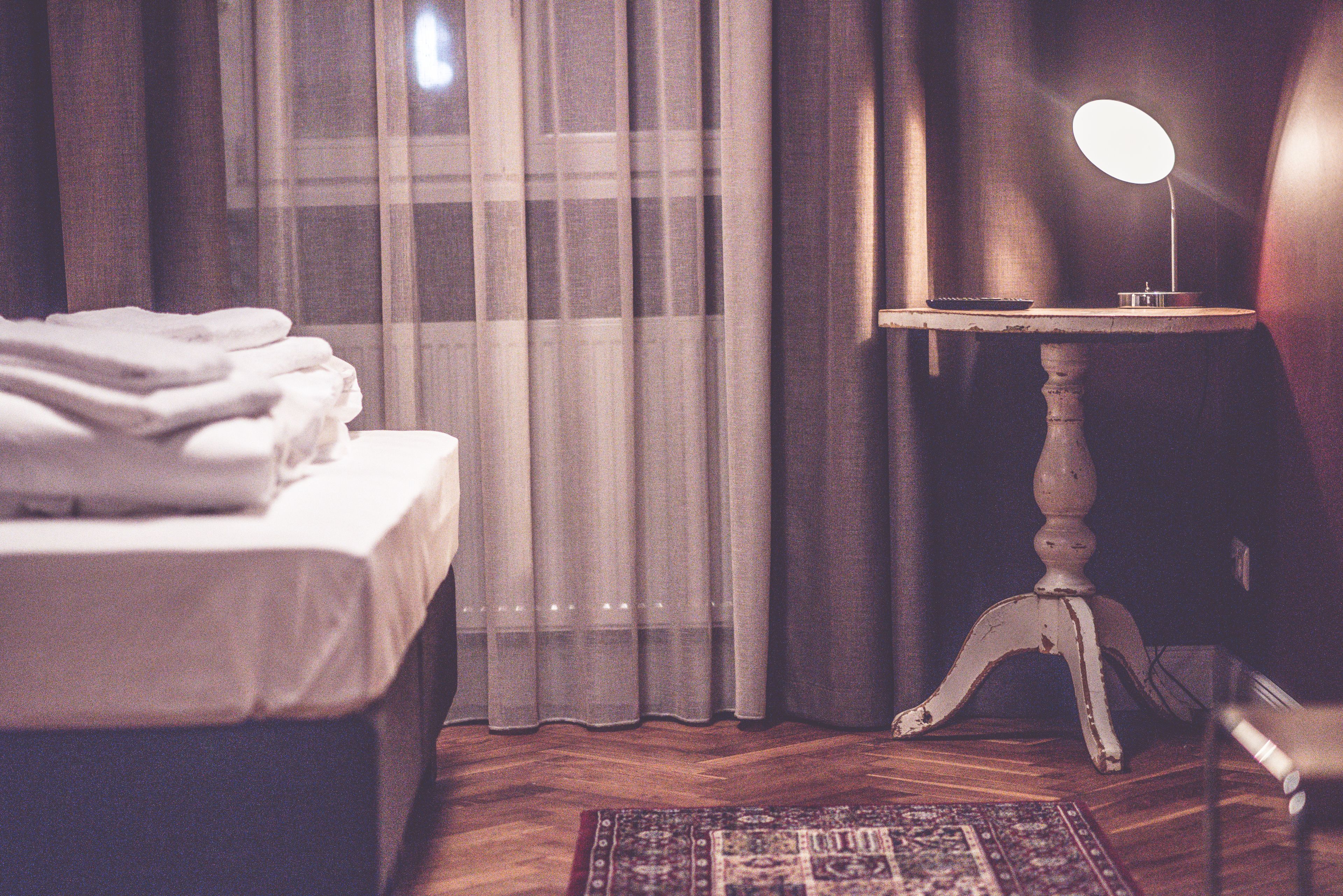 Uma cama bem arrumada com toalhas dobradas, uma mesinha com abajur e um tapete estampado sobre piso de madeira.