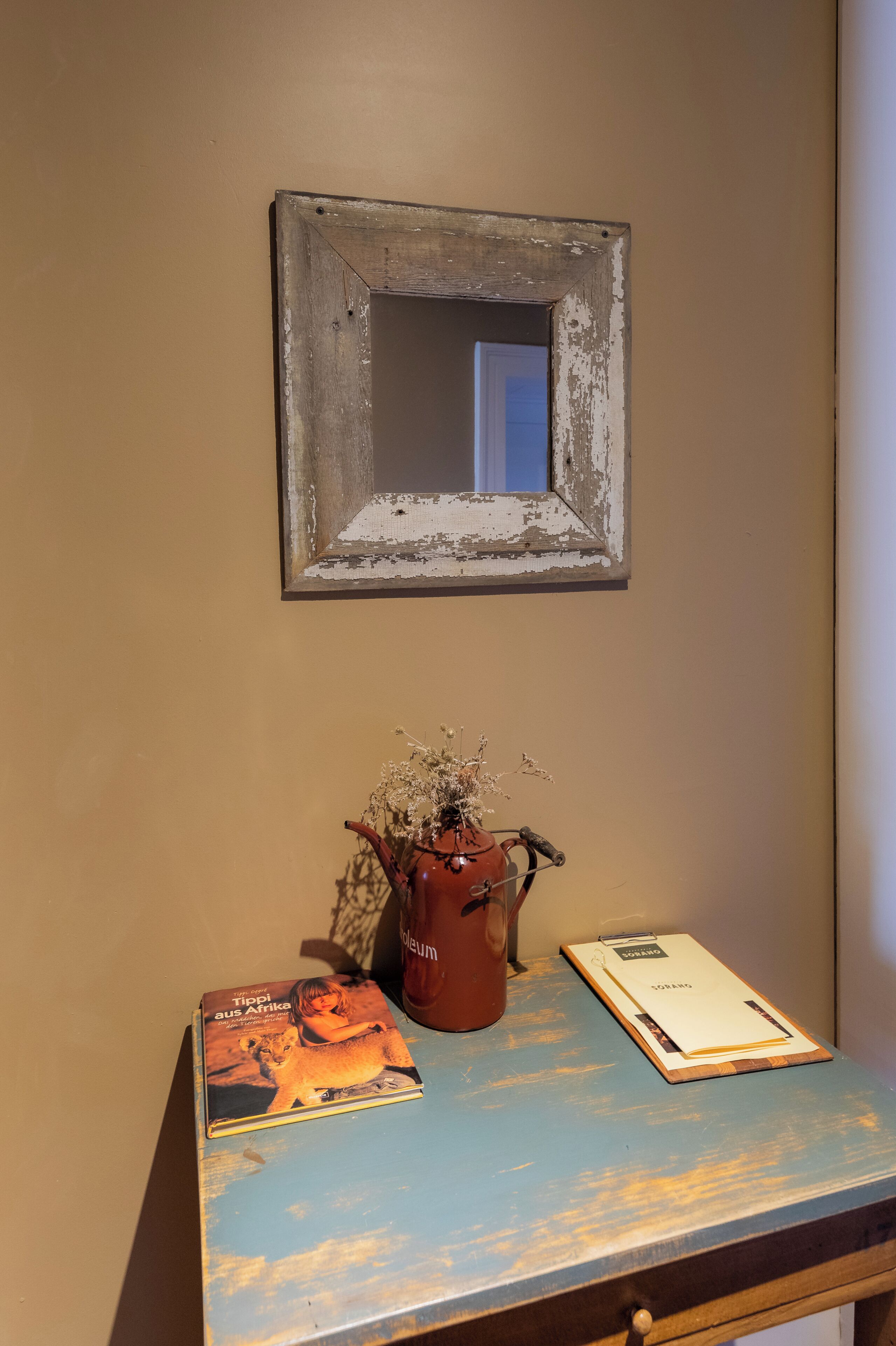 Um espelho fixado na parede, um vaso com flores secas, um livro e um bloco de notas sobre uma mesa azul.