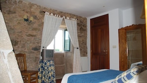 Um quarto com parede de pedra, cama com manta azul, porta de madeira e televisão sobre uma mesa.