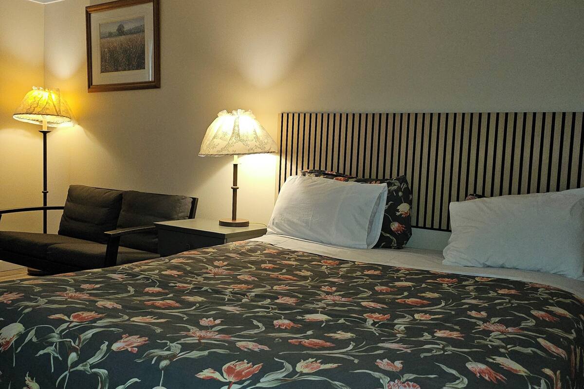 Quarto de hotel com cama grande, duas luminárias de cabeceira, uma poltrona, cabeceira e um quadro na parede.