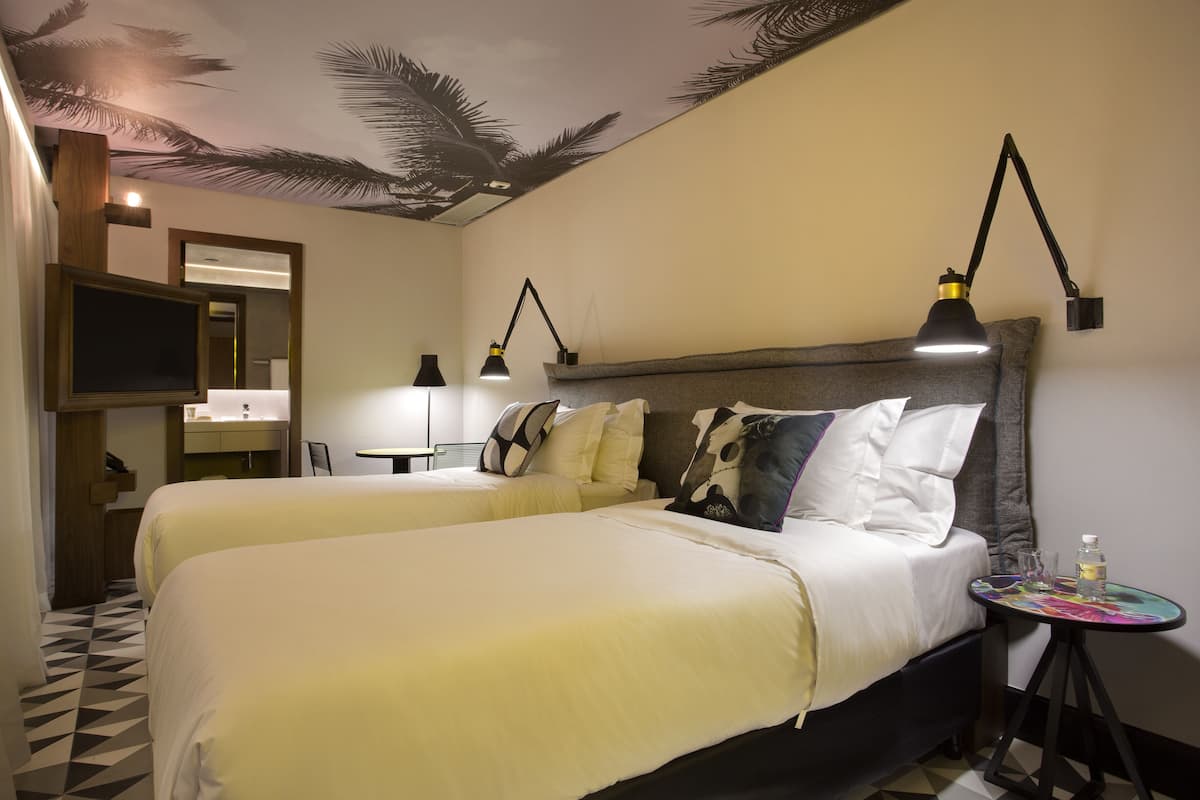 Quarto de hotel com duas camas, uma mesa pequena com abajur, espelho e televisão na parede.