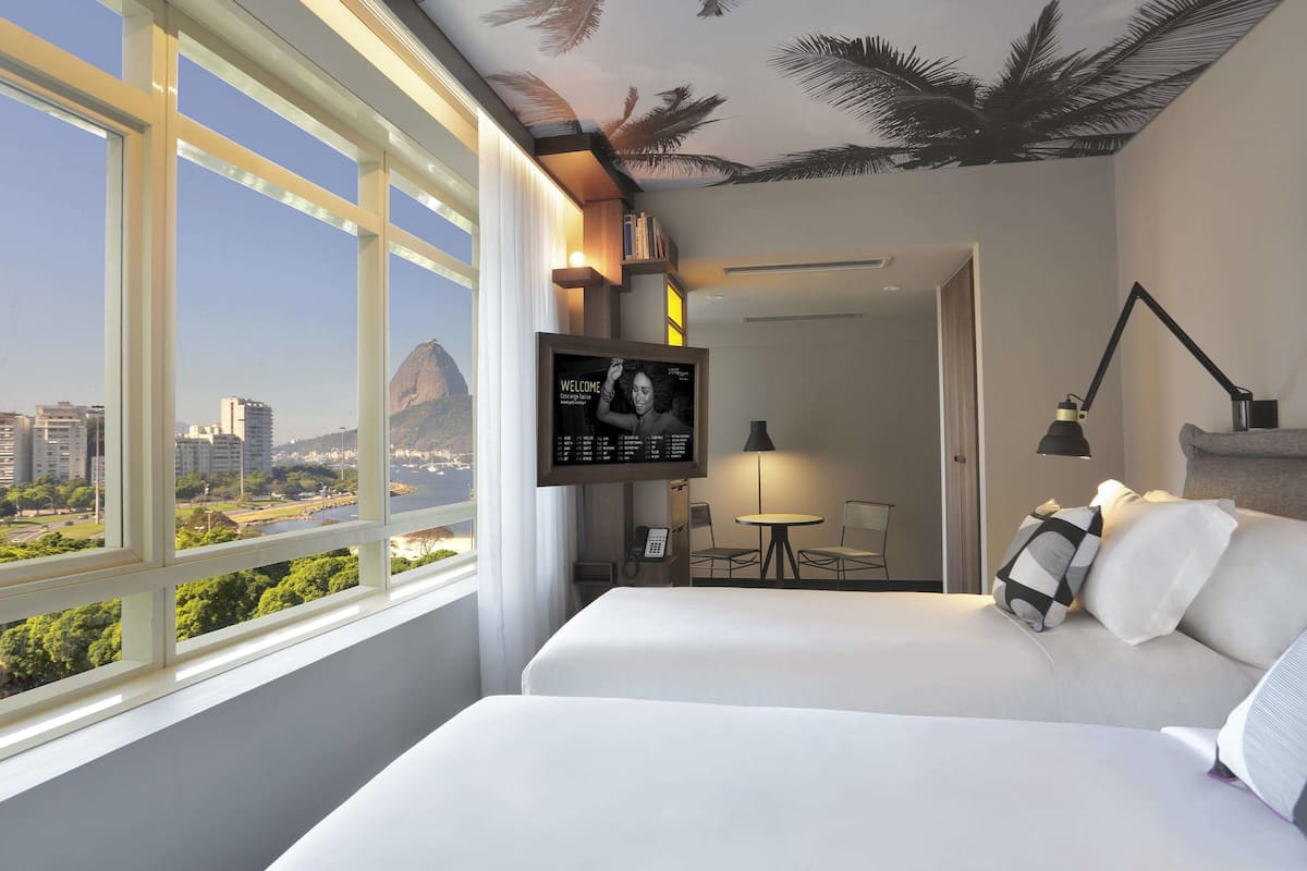 Quarto de hotel com uma grande janela com vista para a cidade, uma cama com almofadas, uma escrivaninha com abajur e uma televisão na parede.