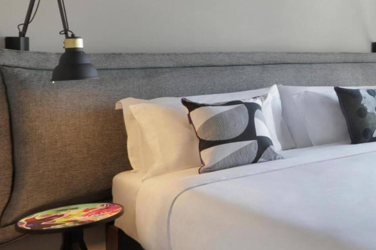 Uma cama bem arrumada com almofadas brancas e estampadas, cabeceira cinza e uma pequena mesa redonda com tampo colorido.
