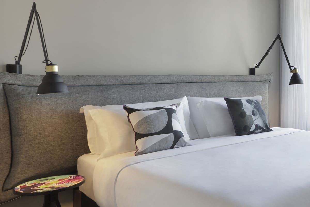 Cama bem arrumada com cabeceira cinza, duas luminárias de mesa pretas e uma mesinha redonda com toalha de mesa colorida.