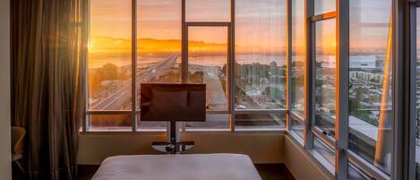 Quarto de hotel com uma cama grande, uma mesa com cadeira e vista para o horizonte urbano ao pôr do sol.