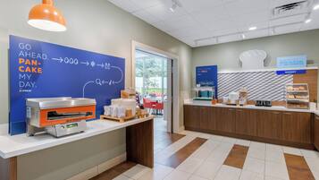 Espaço de café da manhã com TV de tela plana exibindo cardápio, máquina de café e área de padaria com sinalização destacando os favoritos do momento.