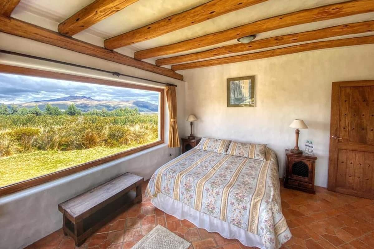 Quarto com uma janela grande, vigas de madeira, uma cama com colcha florida, um banquinho, uma mesinha de cabeceira e um guarda-roupa de madeira.