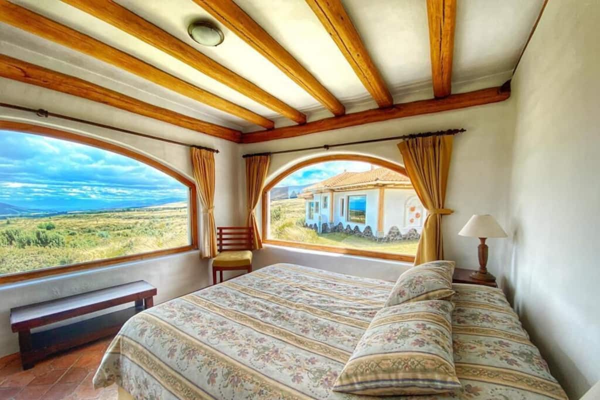Quarto com uma cama grande, vigas de madeira, duas janelas com cortinas, um banco e vista para uma casa e paisagem.