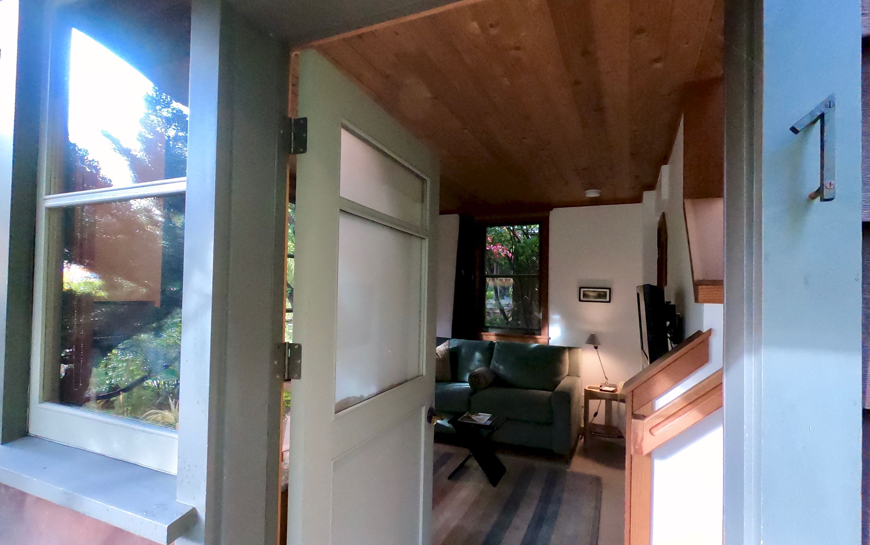 Uma sala de estar com um sofá, uma mesa de centro e uma televisão. Há uma janela com vista para árvores e uma porta que dá para o exterior.