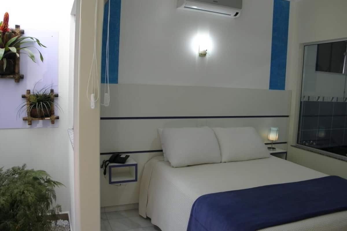 Quarto com cama, mesa de cabeceira, luminária, aparelho de ar condicionado e plantas fixadas na parede.