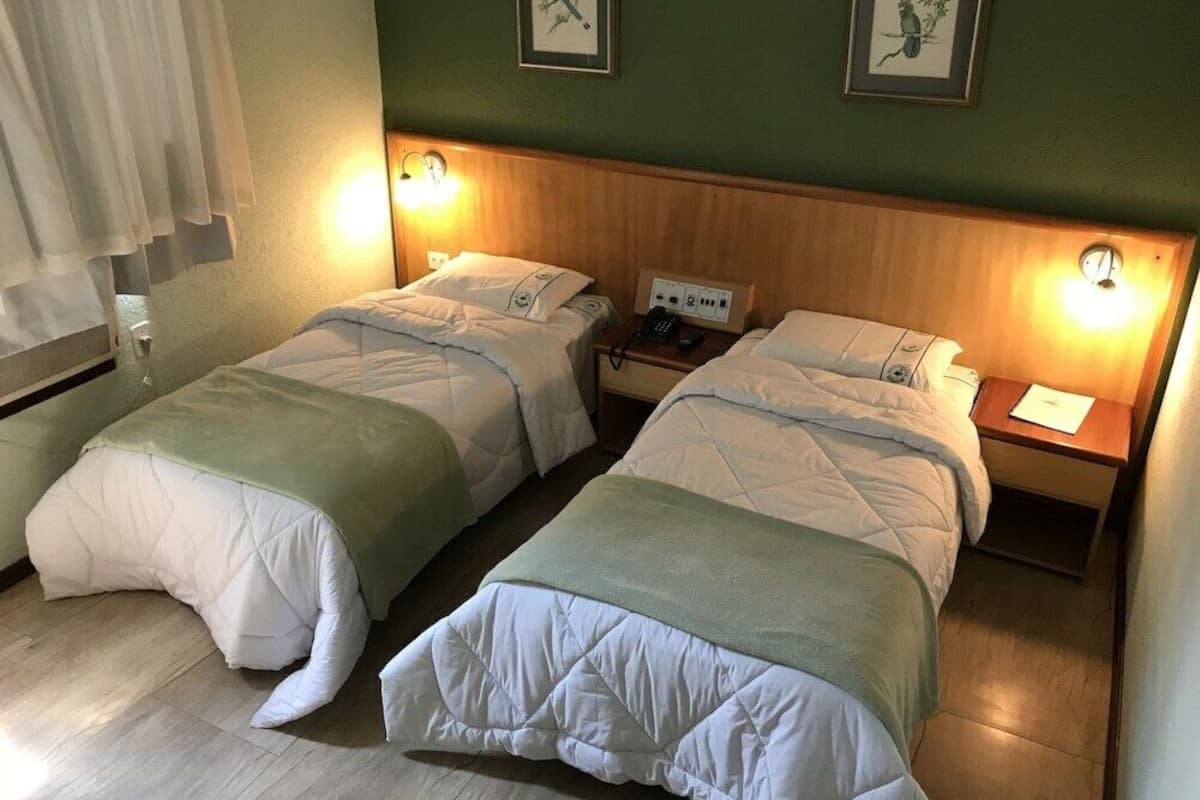 Duas camas de solteiro com cabeceiras, mesas de cabeceira e luminárias fixadas na parede em um quarto com piso de madeira e paredes verdes.