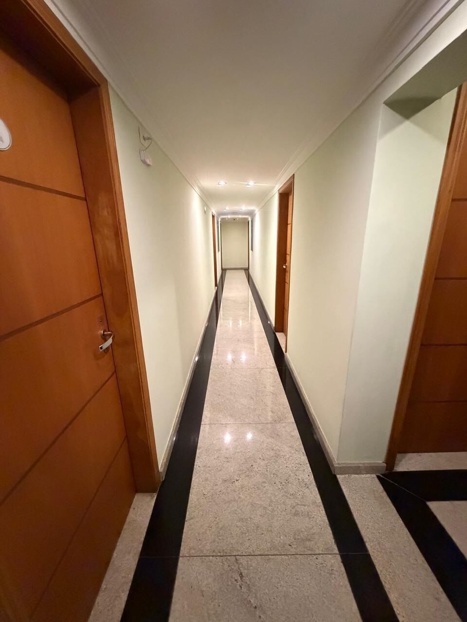 Corredor com portas de madeira em ambos os lados, que dá para um hall com chão de azulejos e iluminação embutida.