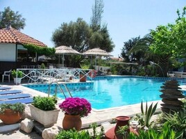 Piscina com espreguiçadeiras e guarda-sóis, cercada por plantas em vasos e um edifício com telhado em azulejos.