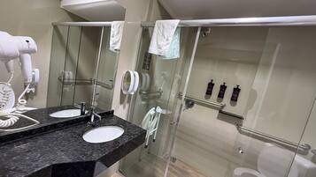 Casa de banho moderna com duas bacias, secador de cabelo, cabine de duche em vidro e artigos de higiene pessoal numa prateleira.