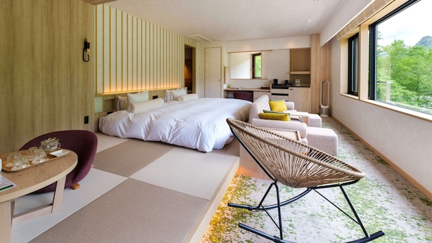 Quarto de hotel moderno com uma cama grande, área de estar com uma poltrona, uma mesinha e vista para o verde lá fora.