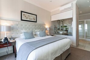 Quarto de hotel moderno com uma cama grande, cabeceira, duas mesas de cabeceira com luminárias, um quadro emoldurado na parede e banheiro com espelho amplo e chuveiro de vidro.