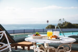 Mesa com itens de café da manhã e um copo de suco de laranja, com vista para uma piscina e o oceano.