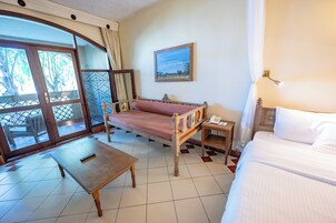 Quarto com cama, mesa, cadeira, mesinha com telefone e janela com vista para árvores.
