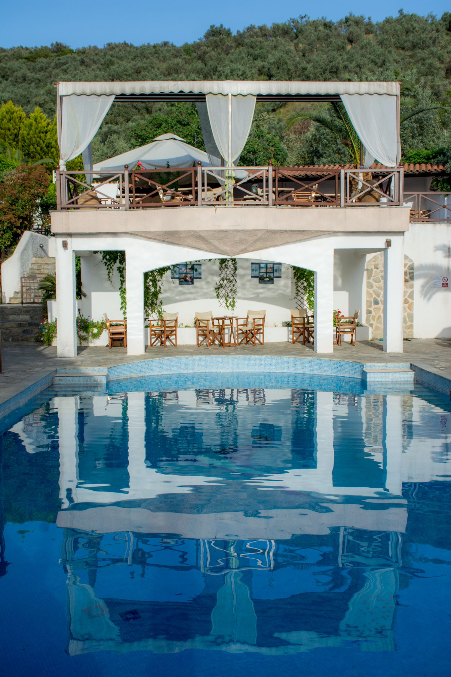 Uma piscina com superfície de água azul cristalina, rodeada por área de estar externa e um espaço coberto com cortinas.
