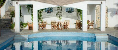 Uma piscina com superfície de água azul cristalina, rodeada por área de estar externa e um espaço coberto com cortinas.