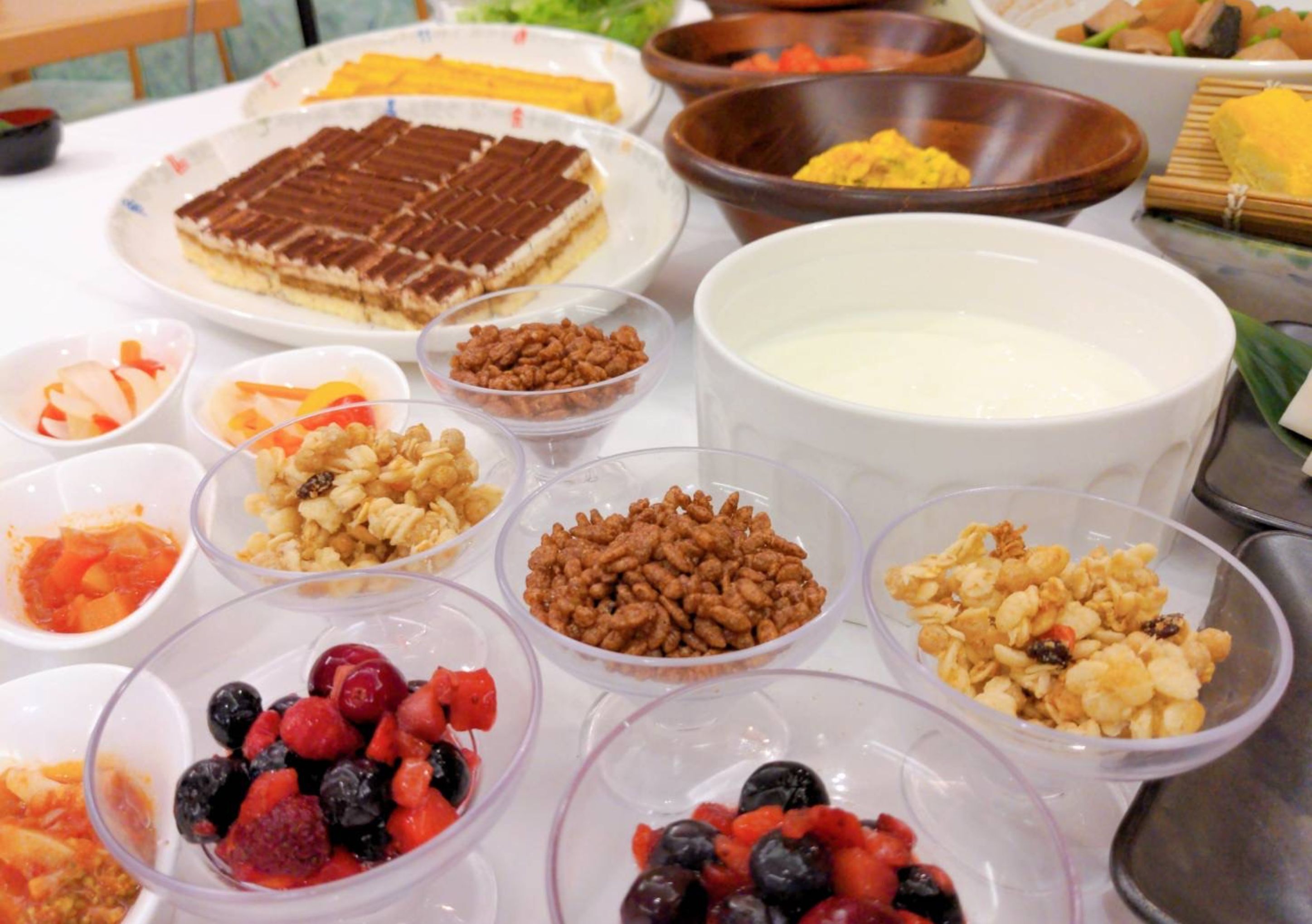 Uma mesa de buffet com diversos alimentos, incluindo sobremesa de chocolate, frutas, iogurte e granola em tigelas.