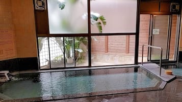 Piscina interior com paredes de vidro transparente, chão em mosaico e área externa visível com plantas.