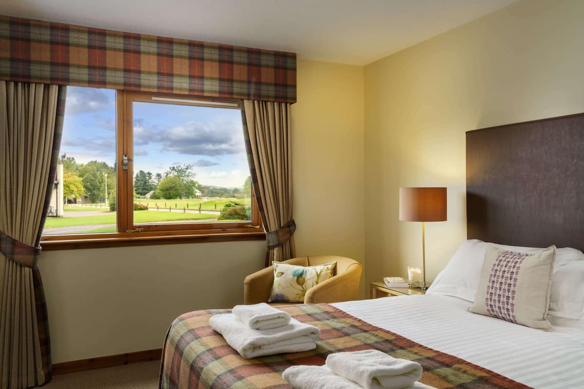 Quarto de hotel com uma cama, uma cadeira, uma luminária e vista para uma paisagem verde pela janela.
