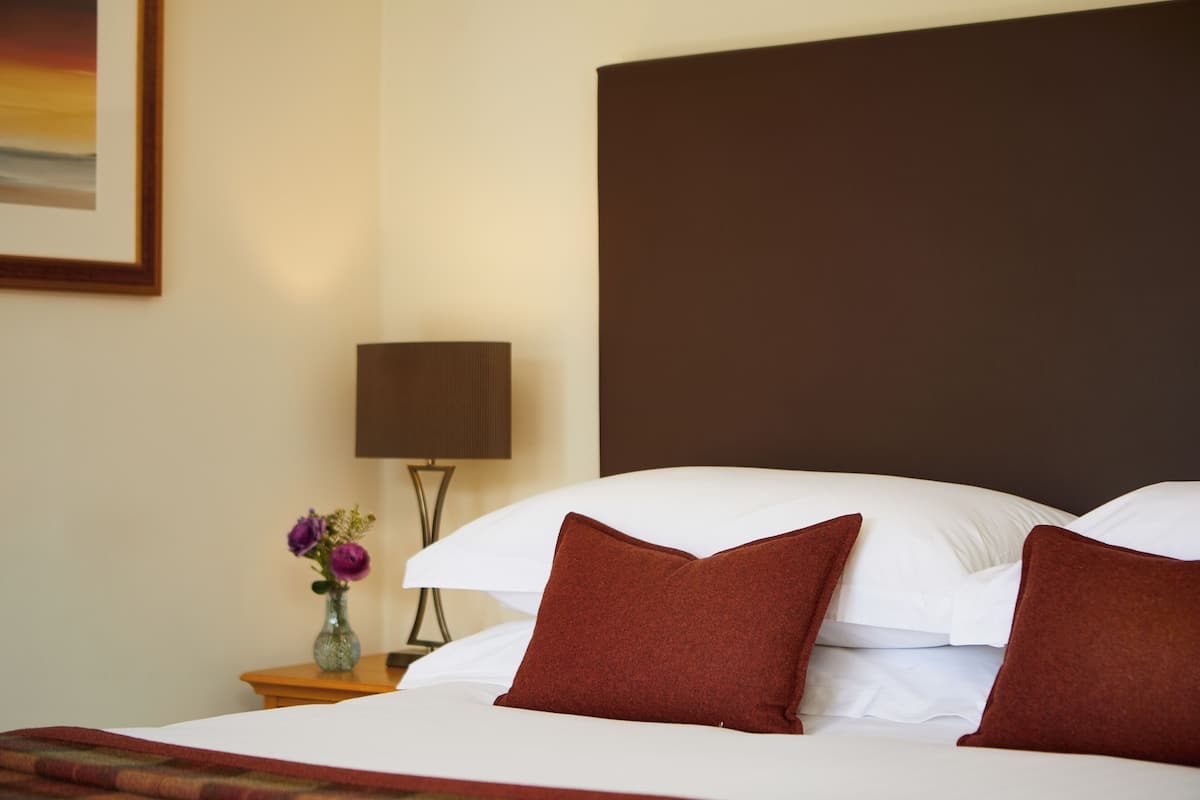 Quarto de hotel com cama, almofadas, um vaso com flores, abajur e um quadro na parede.