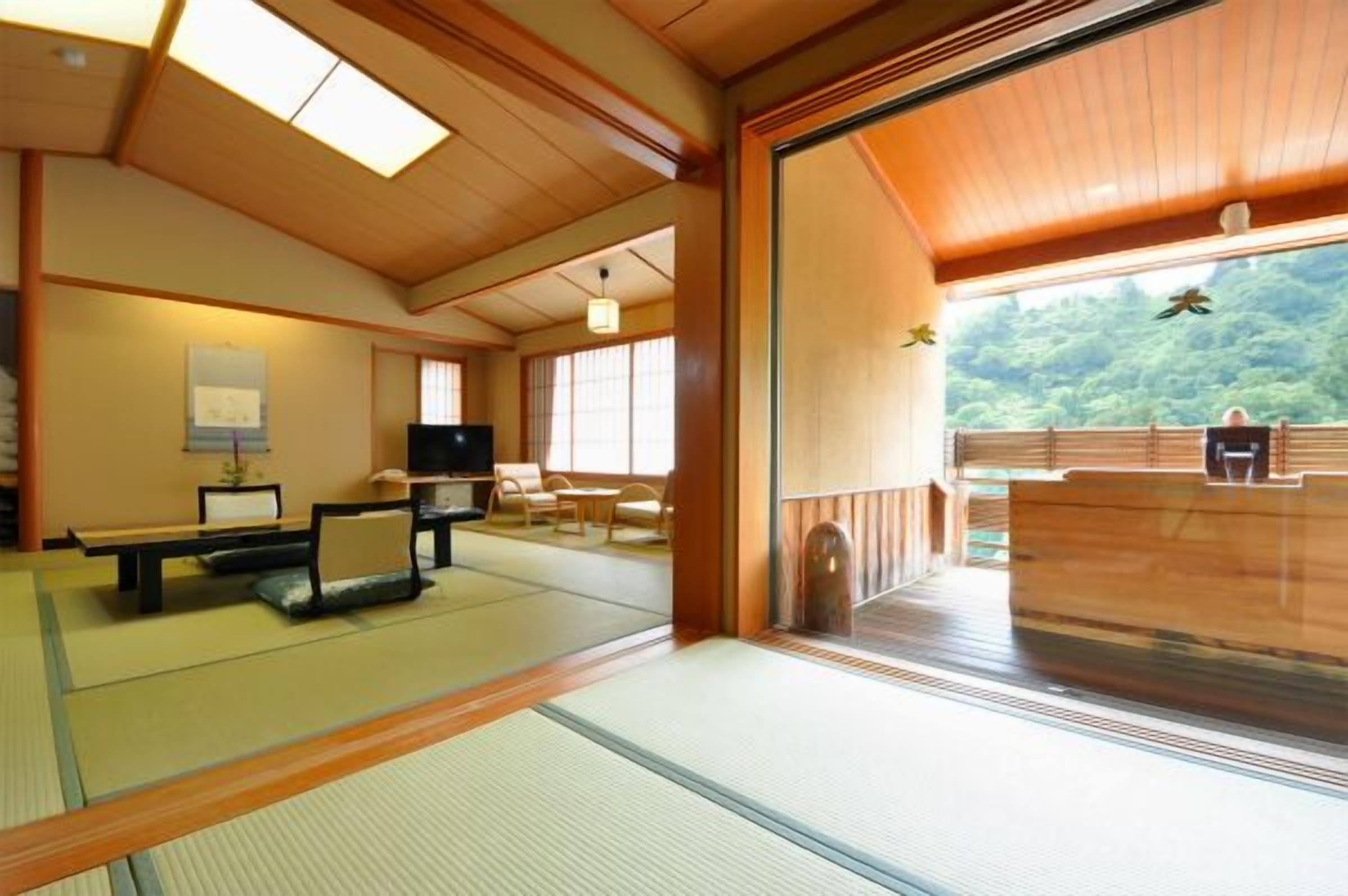 Um quarto japonês tradicional com tatames, uma mesa baixa, televisão e vista para uma área arborizada.