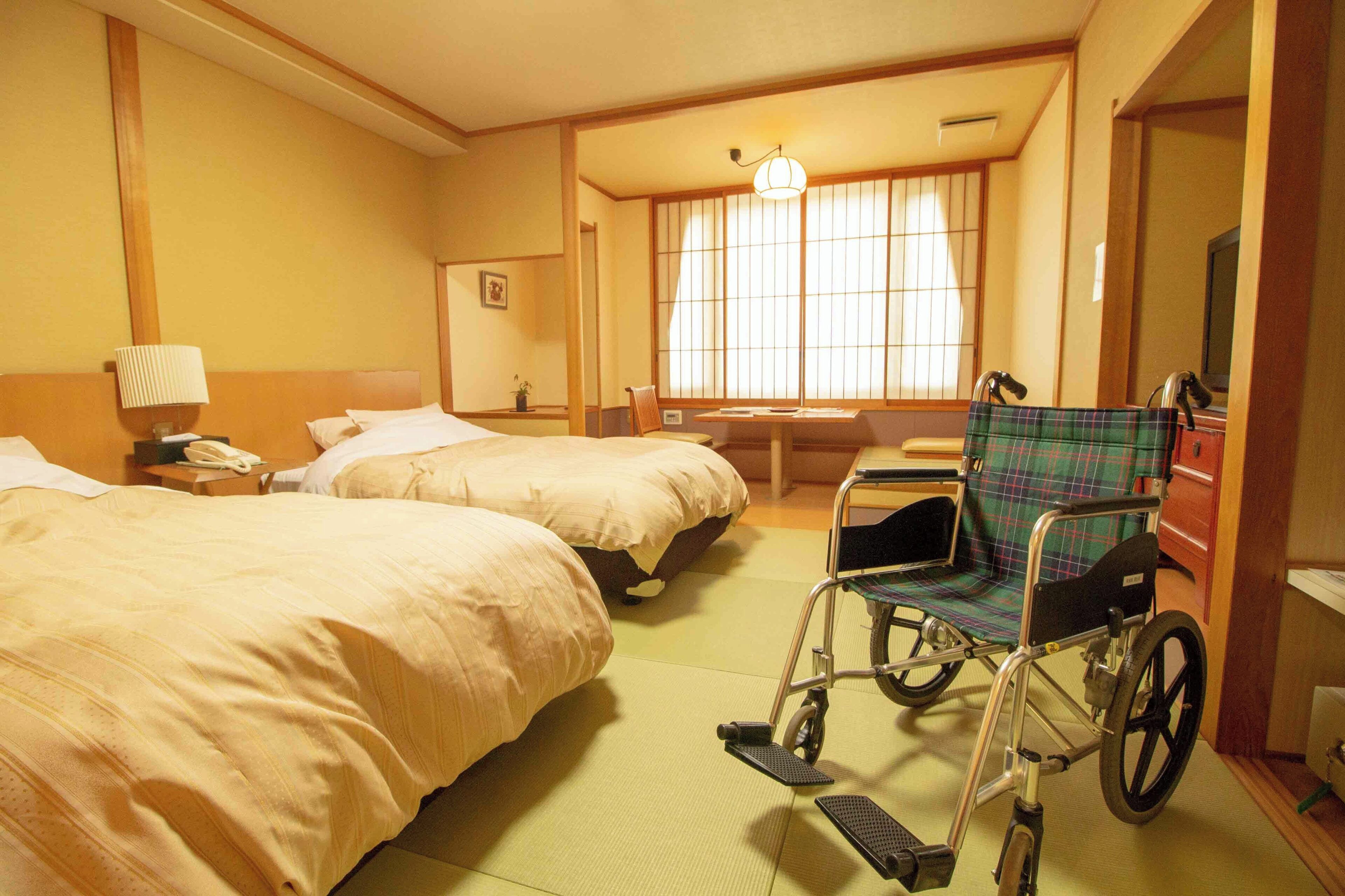 Quarto de hotel com duas camas, uma cadeira de rodas, mesa de cabeceira, abajur e janela com cortinas.