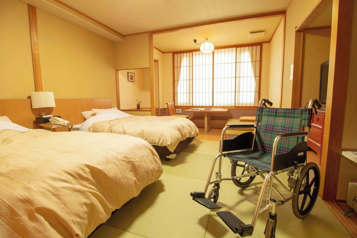 Quarto de hotel com duas camas, uma cadeira de rodas, mesa de cabeceira, abajur e janela com cortinas.