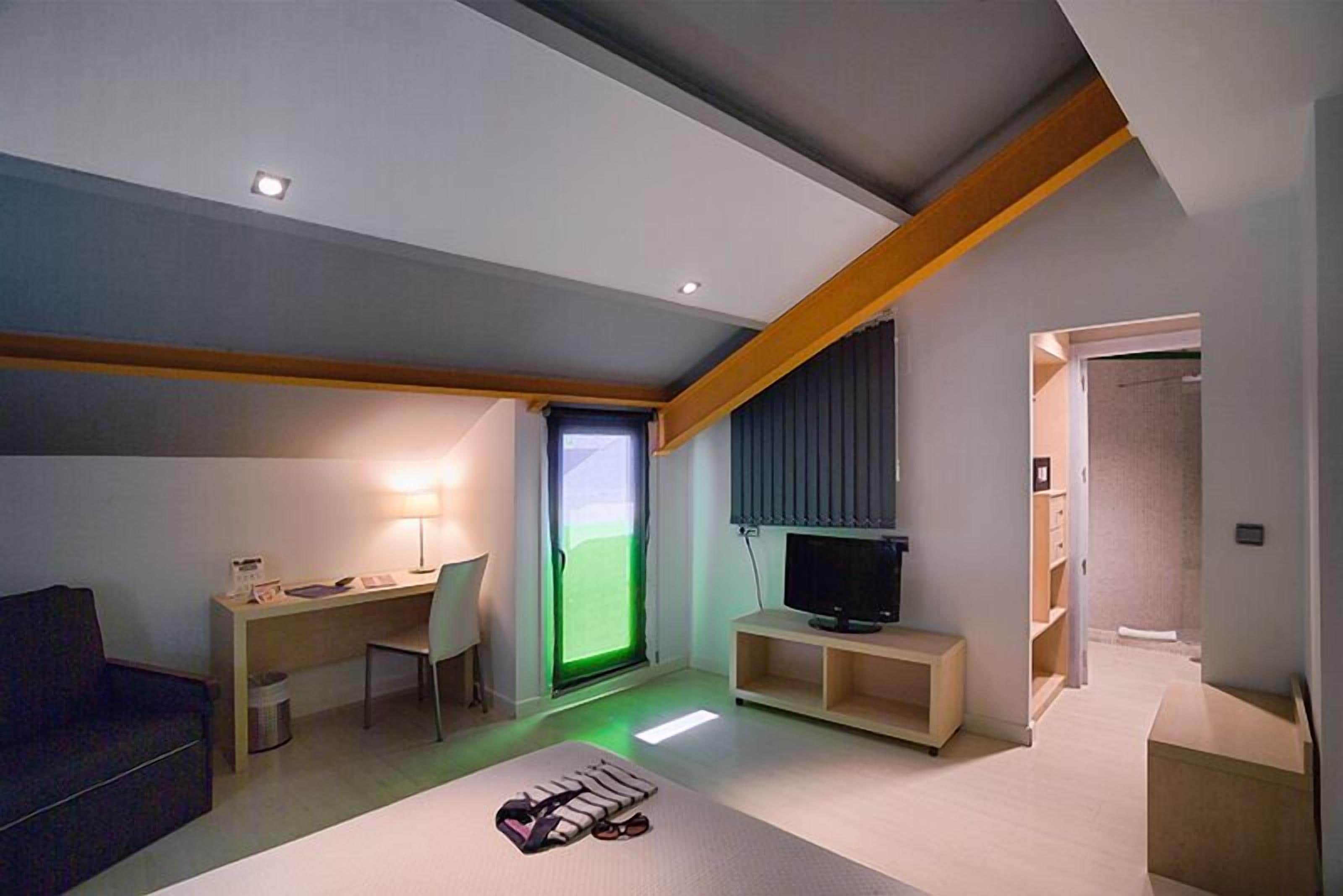 Quarto de hotel moderno com escrivaninha, cadeira, TV e cama.