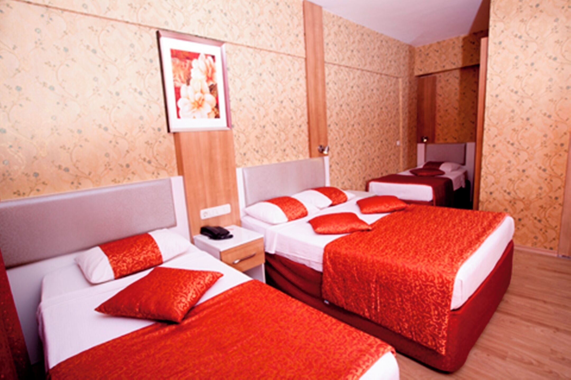 Quarto de hotel com três camas, cada uma com roupa de cama vermelha e almofadas brancas, uma mesa de cabeceira com telefone, e um quadro na parede.