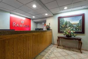 Uma área de recepção com um letreiro vermelho 'RAMADA', um balcão de madeira, piso de azulejos e uma pintura emoldurada de uma paisagem urbana.