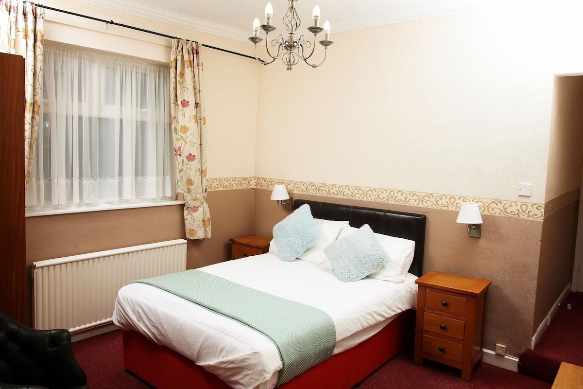 Quarto de hotel com cama, duas mesas de cabeceira, radiador, candelabro e janela com cortinas.