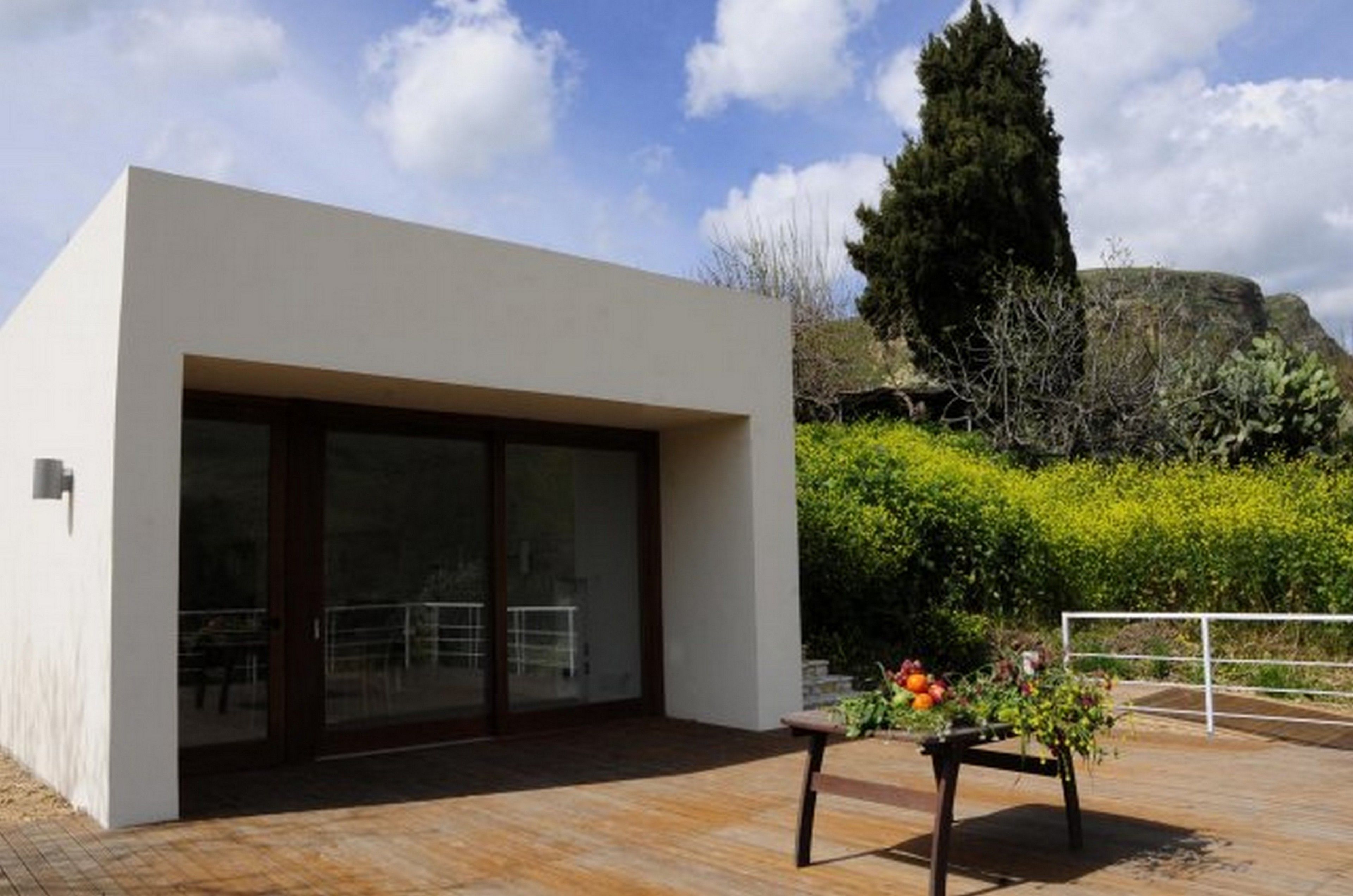 Um edifício moderno com uma grande porta de vidro, área de pátio com mesa e plantas, e vista para uma colina arborizada.