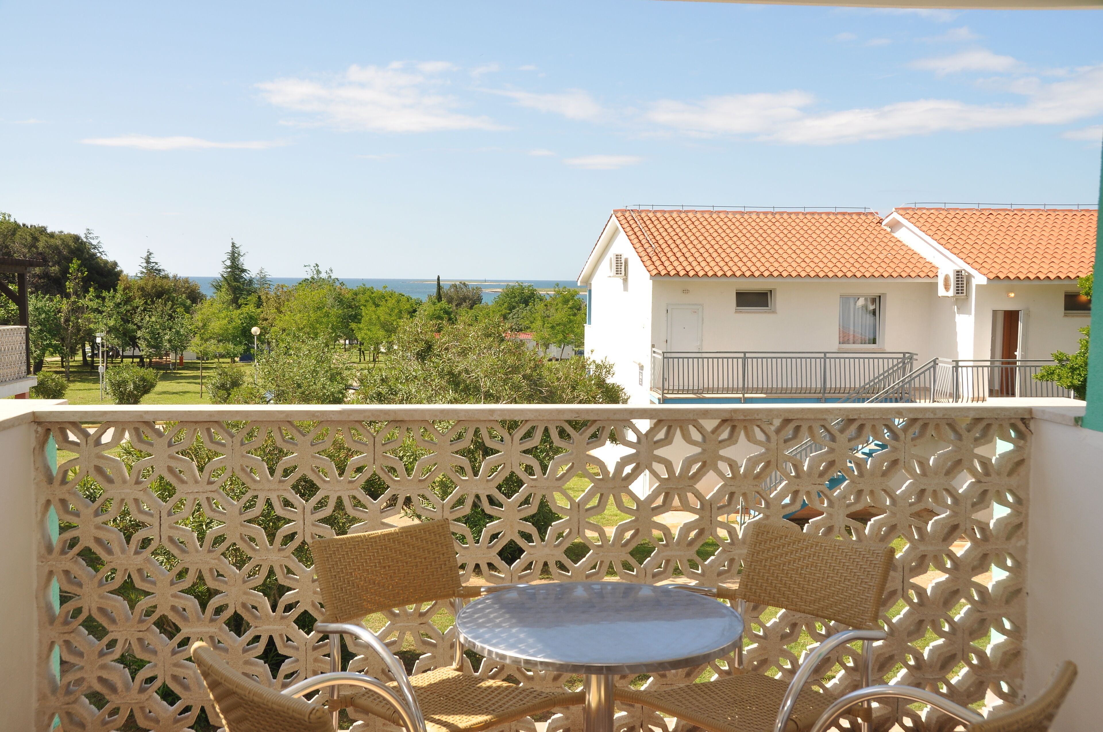 Varanda com cadeiras de vime e uma mesa redonda, com vista para um edifício branco com telhado vermelho e uma piscina.