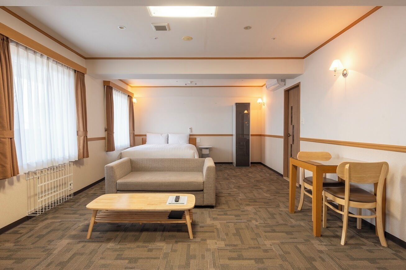 Quarto de hotel com uma cama, um sofá, uma mesinha e uma cadeira. Há um radiador na parede e uma porta ao fundo.