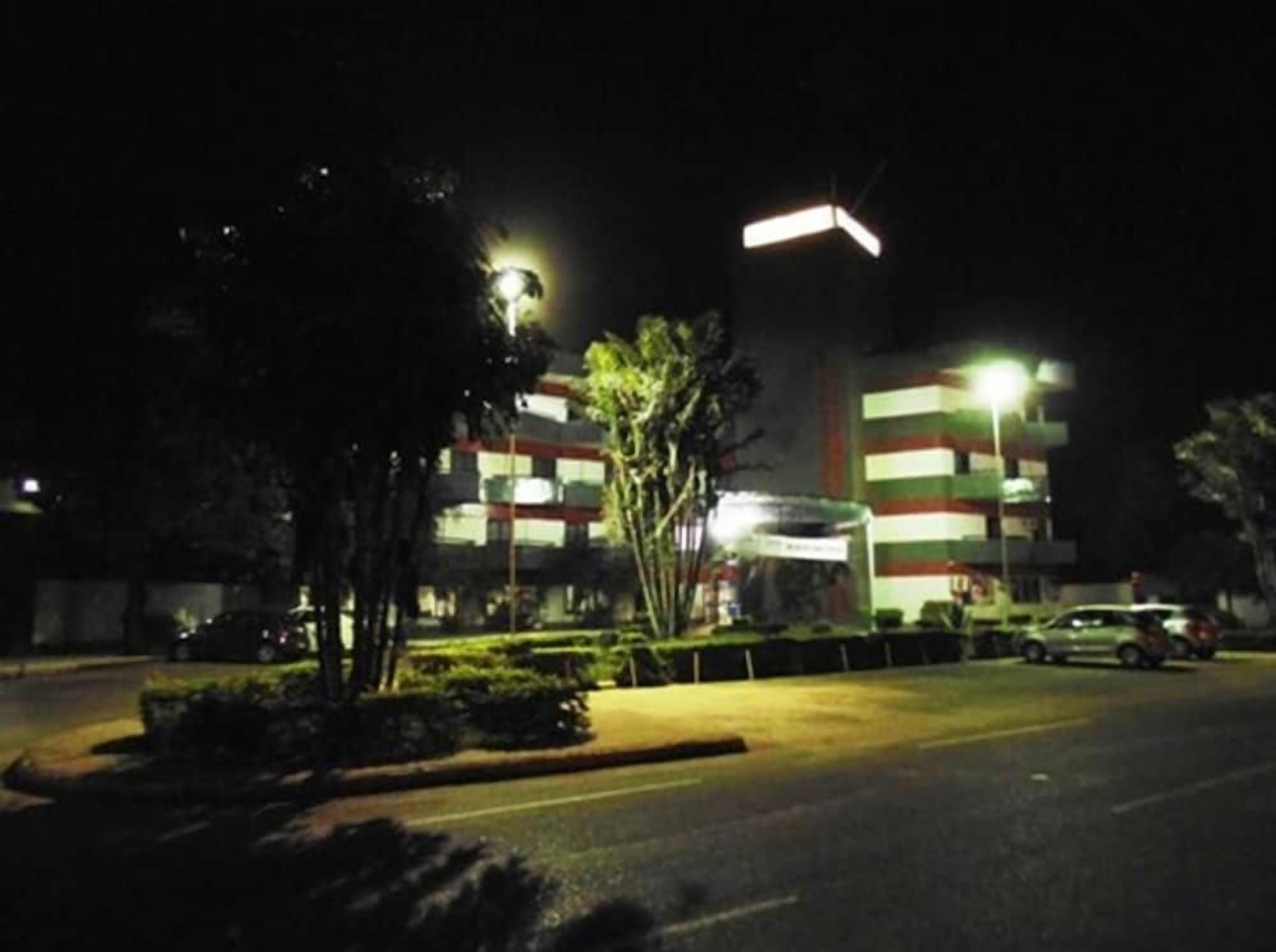 Um edifício com vários andares, iluminado por postes de luz, cercado por uma área de estacionamento.