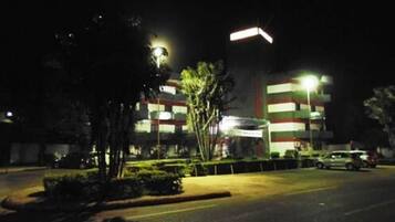 Um edifício com vários andares, iluminado por postes de luz, cercado por uma área de estacionamento.