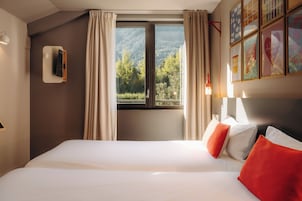 Quarto de hotel com uma cama grande, janela com cortinas, luminária na parede e quadros emoldurados.