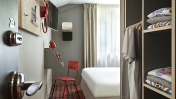 Quarto de hotel com cama, uma cadeira vermelha, escrivaninha com computador, espelho e guarda-roupa.